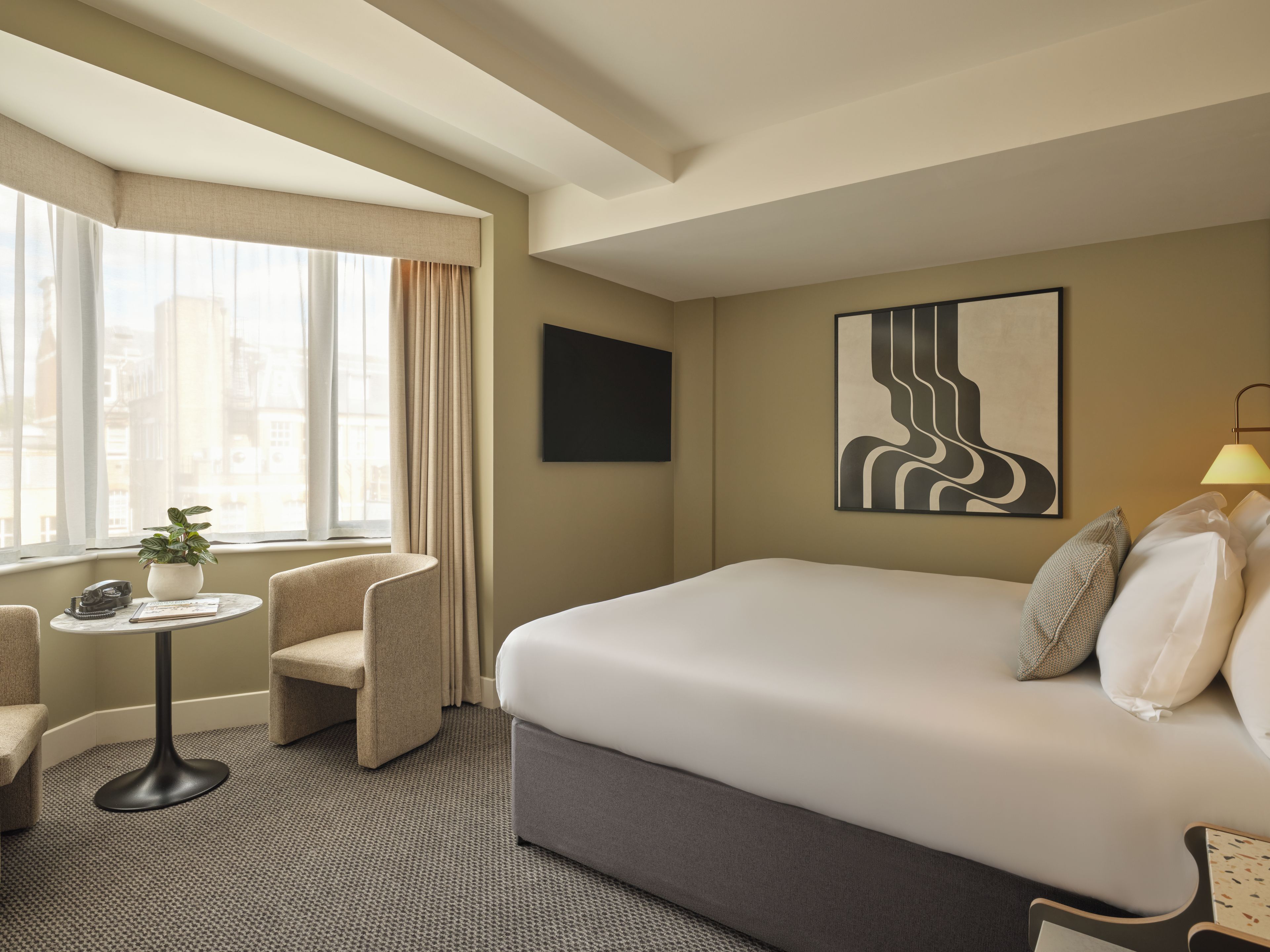 Quarto de hotel com cama, escrivaninha, cadeira, luminária e janela com cortinas.