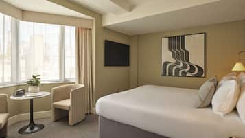 Quarto de hotel com cama, escrivaninha, cadeira, luminária e janela com cortinas.