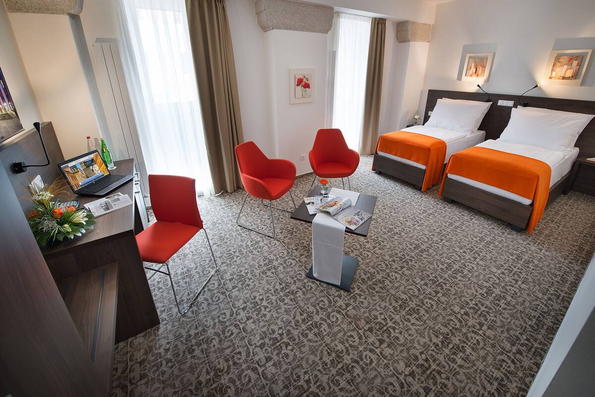 Quarto de hotel com duas camas, uma escrivaninha com um laptop, uma cadeira vermelha e uma mesa de centro com revistas.