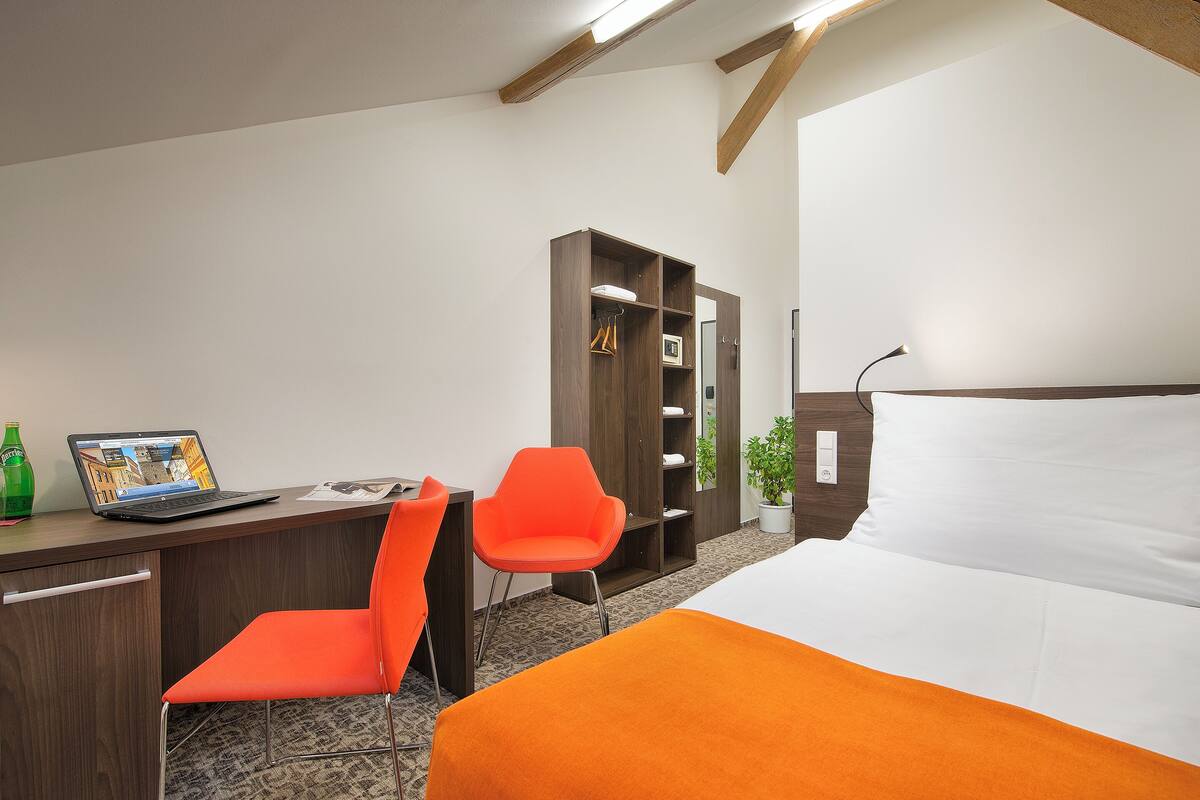Quarto de hotel com uma cama, uma escrivaninha com um laptop, uma cadeira, um guarda-roupa e uma pequena planta em vaso.