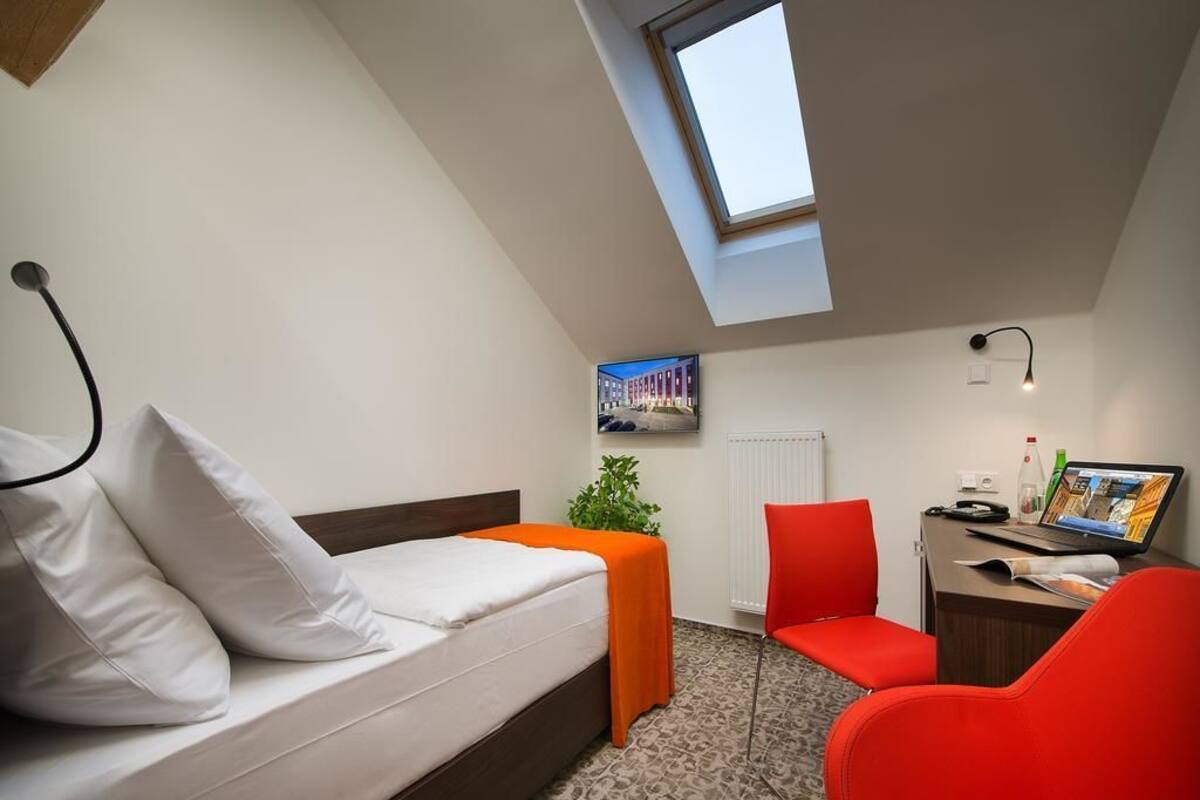 Quarto de hotel moderno com cama, escrivaninha, poltrona, televisão e claraboia.