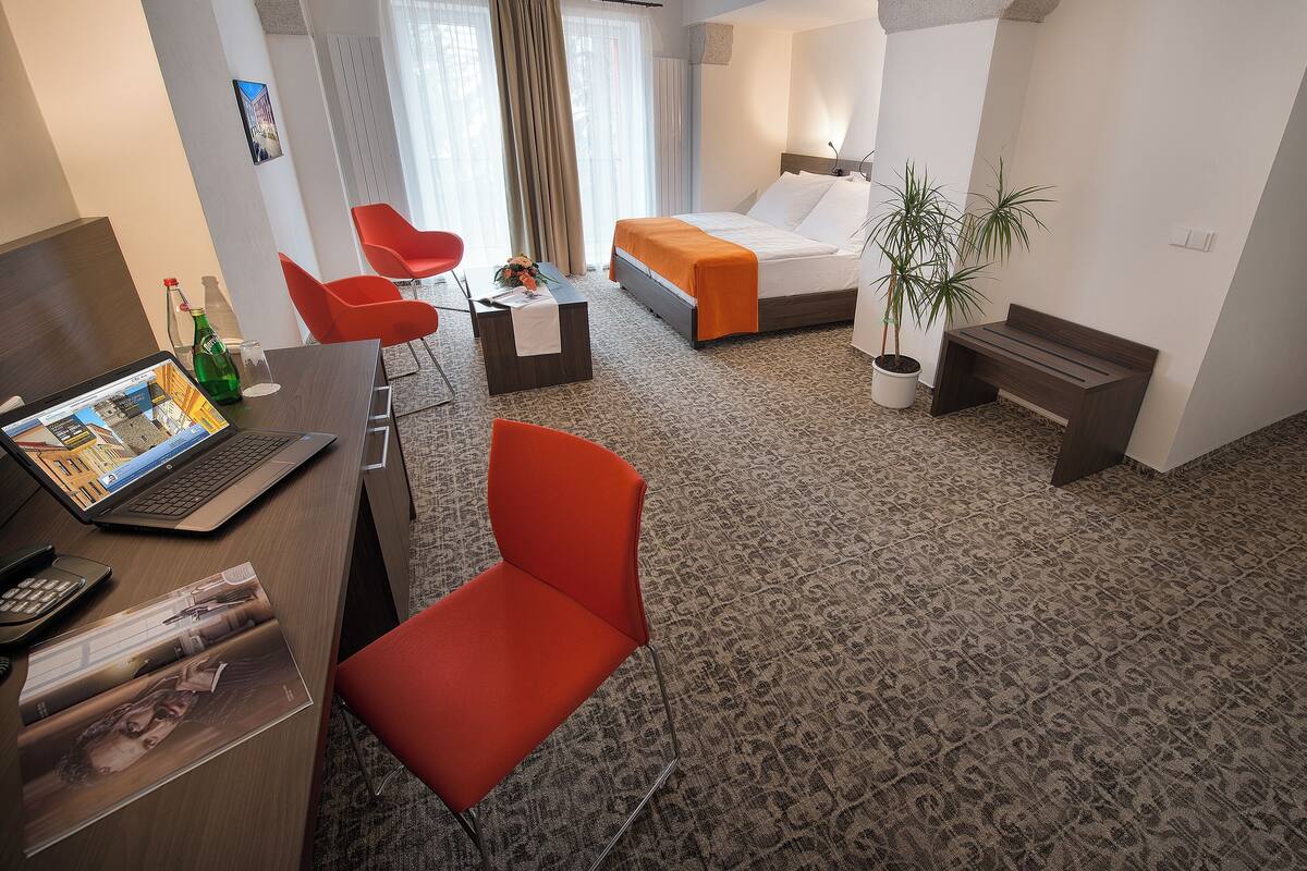 Quarto de hotel com cama, escrivaninha com laptop, cadeira, pequena mesa com planta e janela com cortinas.