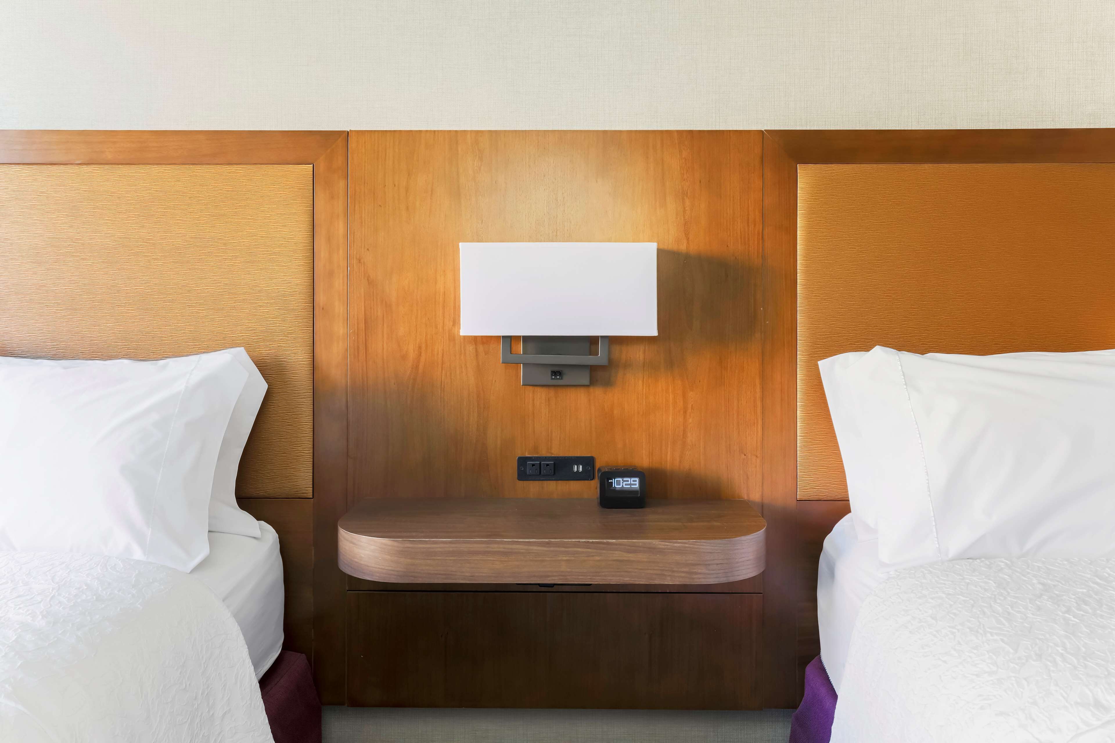 Quarto de hotel com duas camas, uma cabeceira de madeira, uma mesa de cabeceira, uma luminária na parede e um relógio digital.