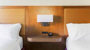 Quarto de hotel com duas camas, uma cabeceira de madeira, uma mesa de cabeceira, uma luminária na parede e um relógio digital.