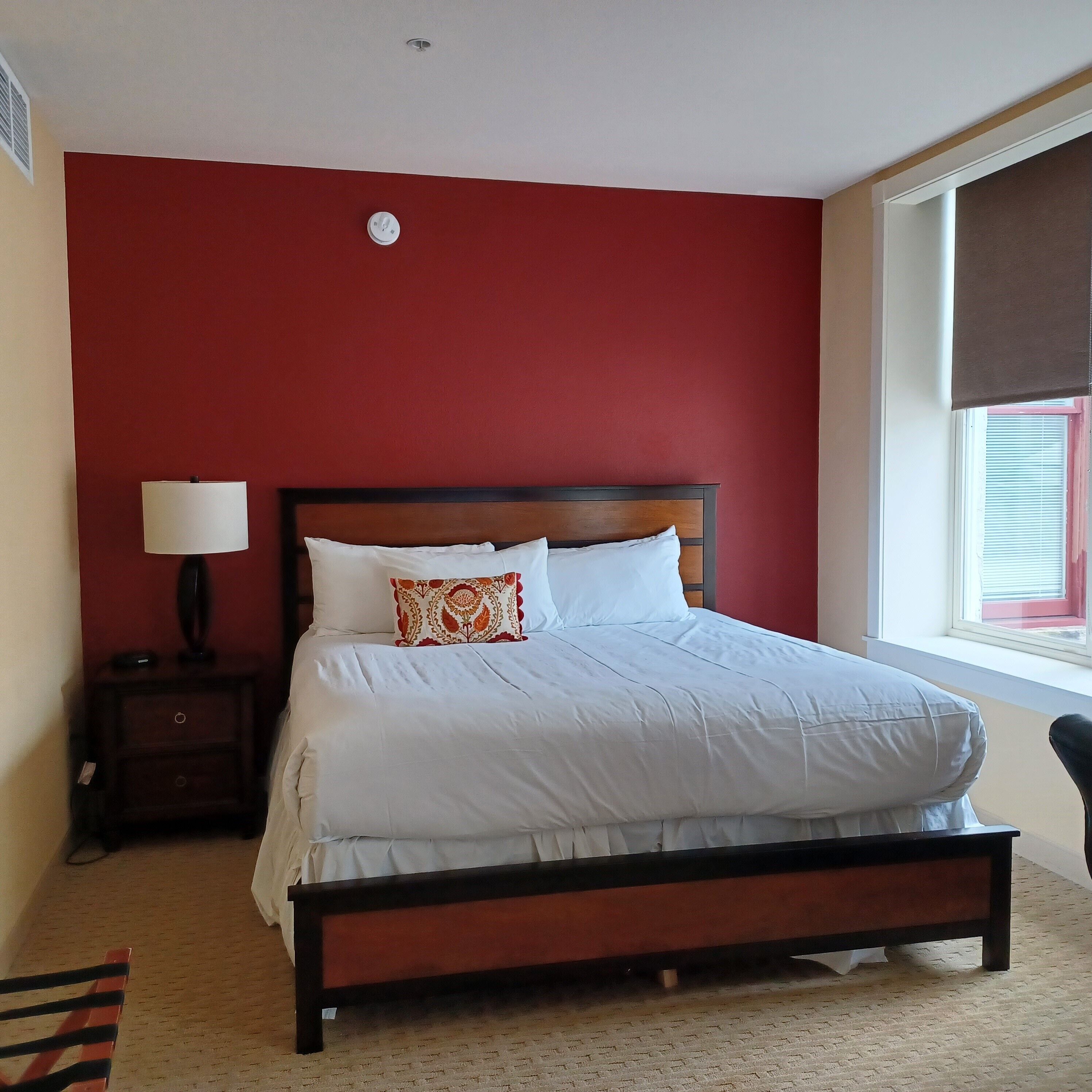 Quarto com cama de madeira, roupa de cama branca, parede de destaque vermelha, abajur e janela com persianas.