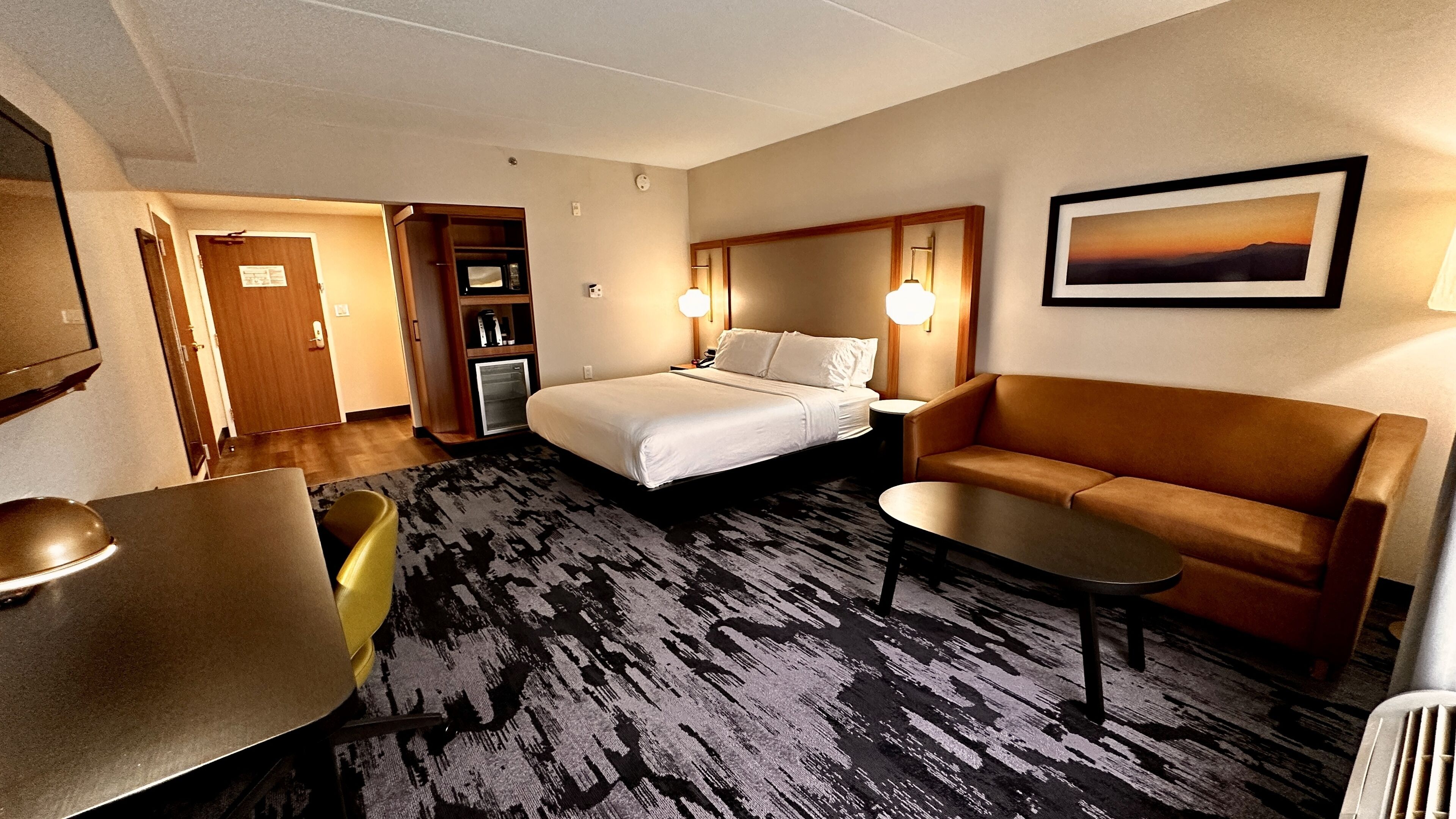 Quarto de hotel com cama, sofá, mesinha, escrivaninha, cadeira, televisão, estante de livros e um quadro na parede.