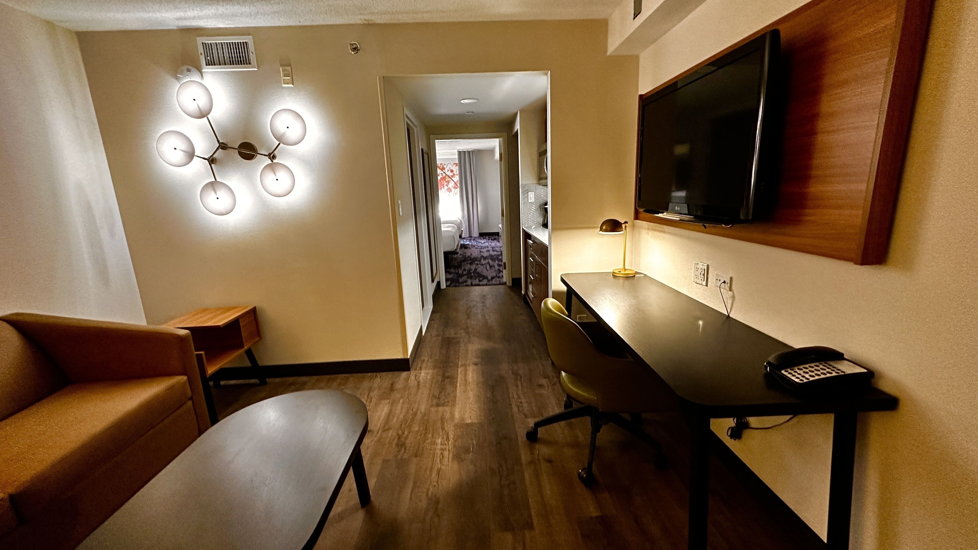 Quarto de hotel com sofá, escrivaninha, TV fixada na parede e luminária no teto.