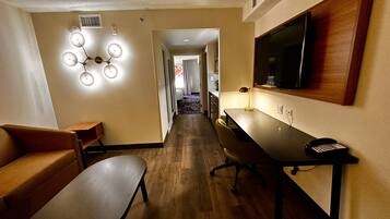 Quarto de hotel com sofá, escrivaninha, TV fixada na parede e luminária no teto.