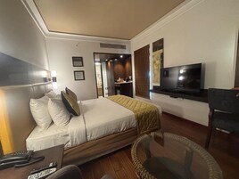 Quarto de hotel com cama, televisão, cadeira, secretária com telefone e controles remotos, e banheiro visível por uma porta aberta.