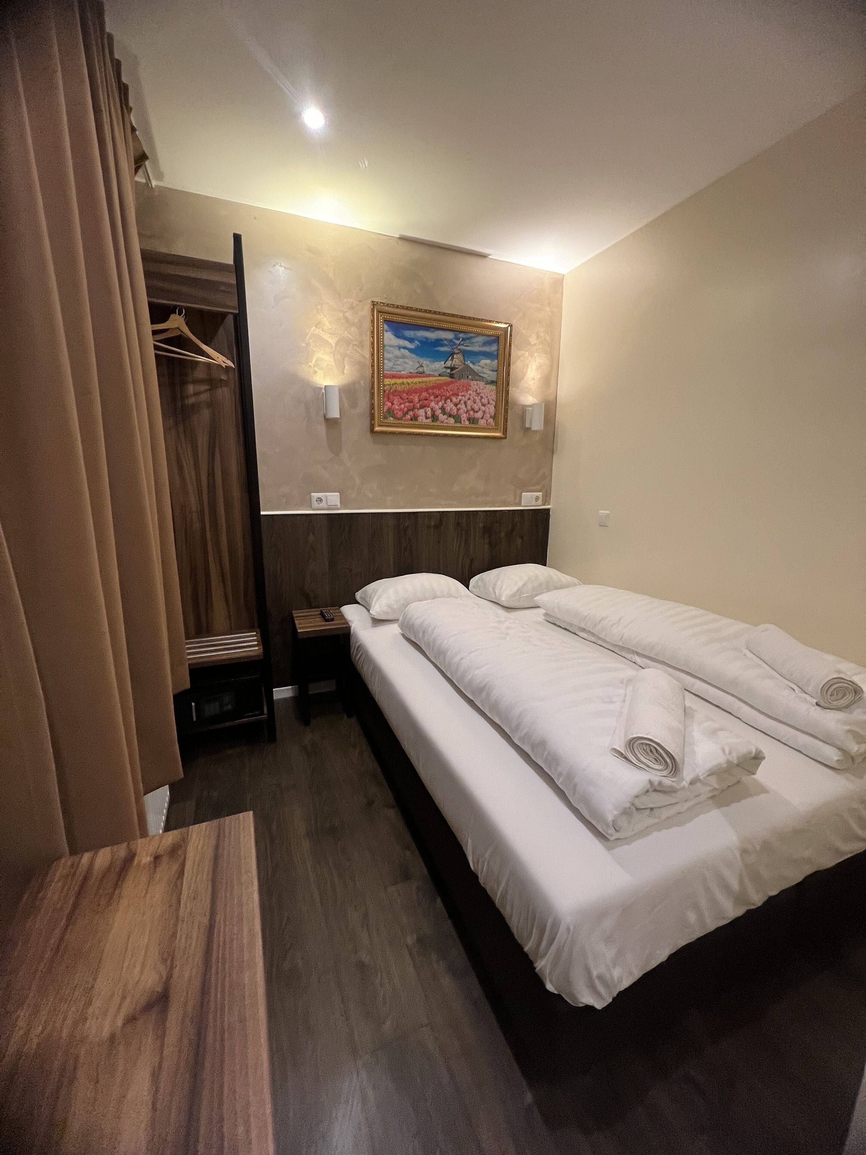 Quarto de hotel com cama, mesa de cabeceira de madeira, escrivaninha, cabideiro e um quadro na parede.