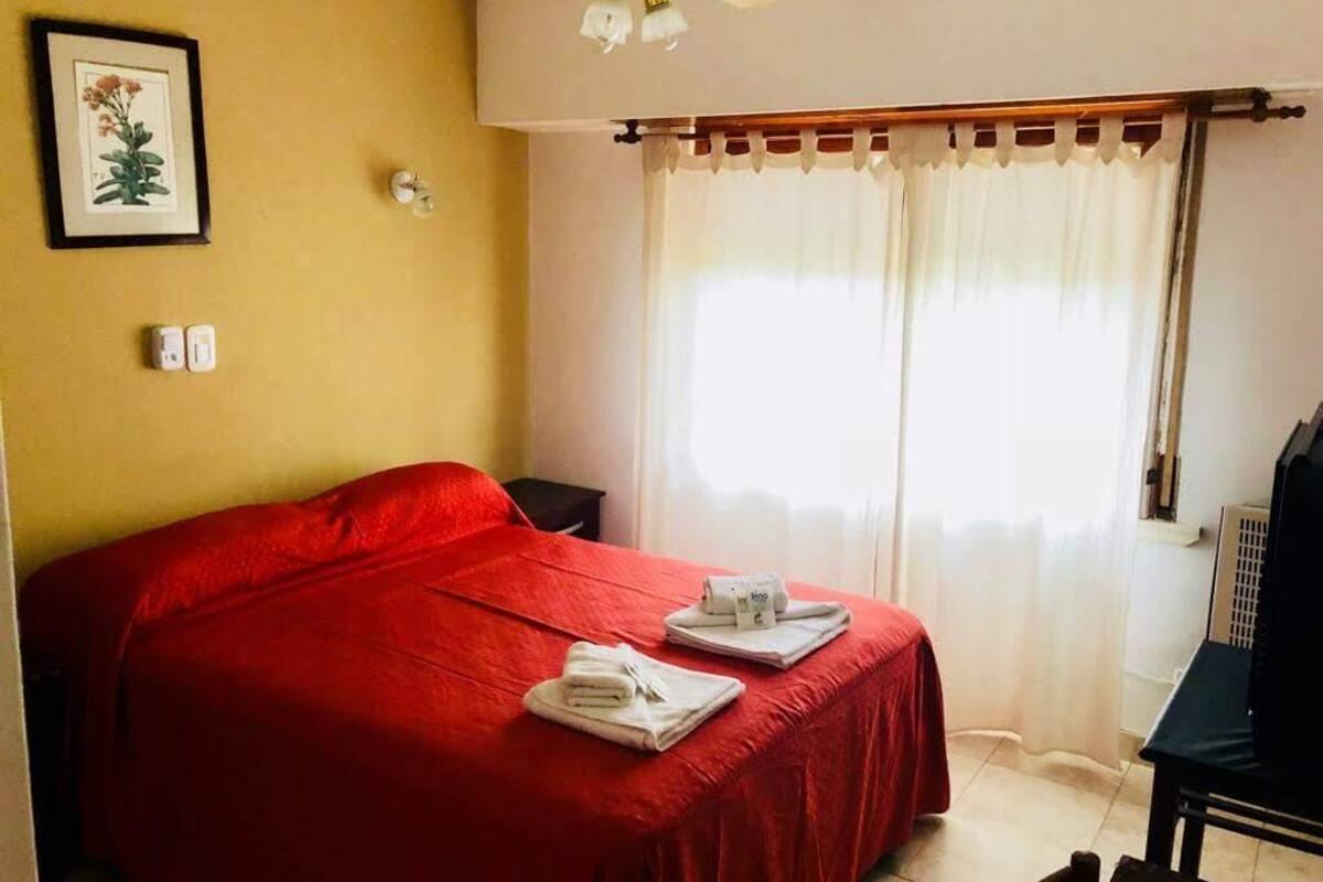 Quarto com cama, ventilador de teto, janela com cortinas, quadro pendurado na parede e uma cadeira.