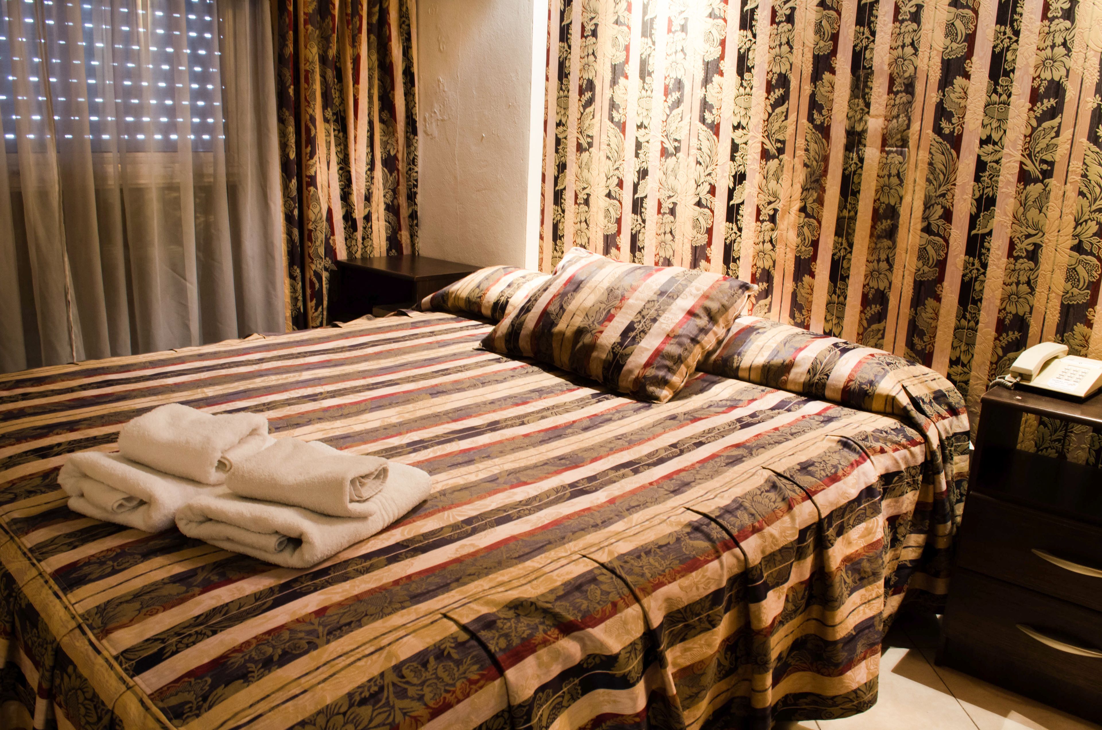 Uma cama bem arrumada com roupa de cama listrada e duas almofadas, uma mesa de cabeceira com telefone, e cortinas com padrão floral.
