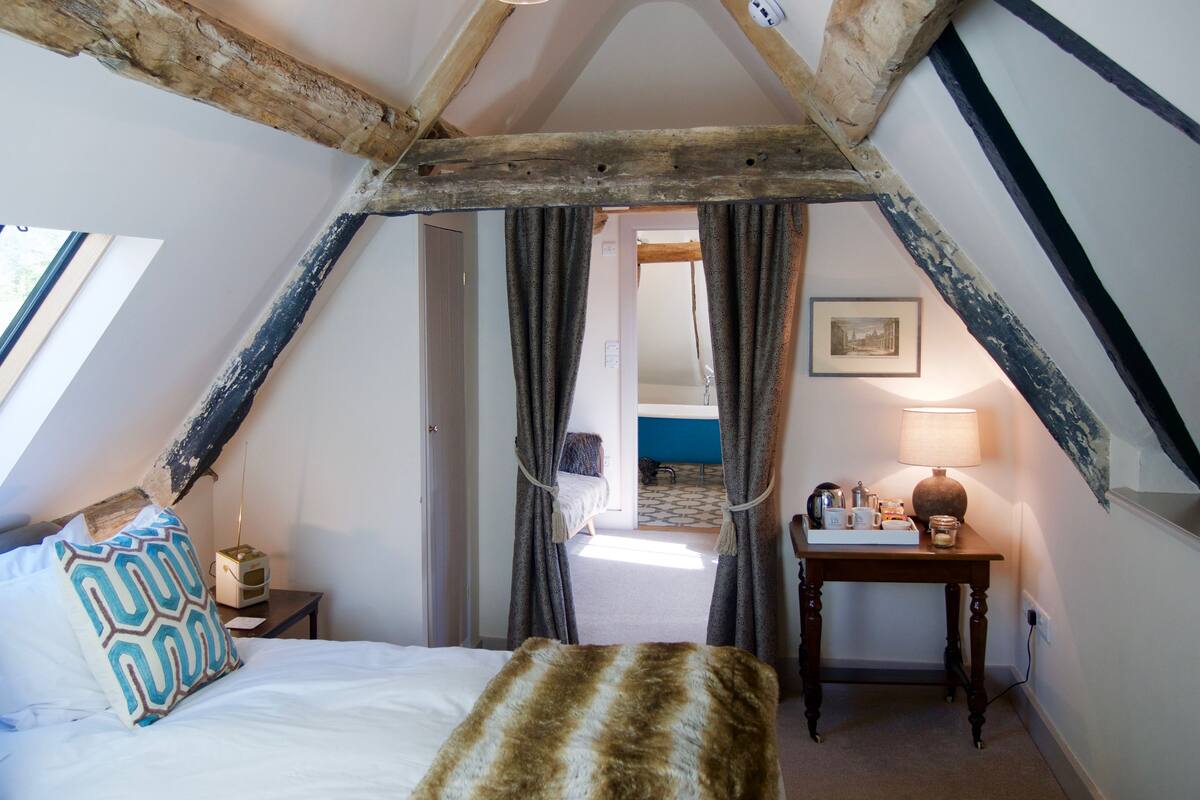 Um quarto de sótão acolhedor, com vigas de madeira expostas, uma cama com almofadas estampadas, uma mesa de cabeceira com abajur e vista para o banheiro.