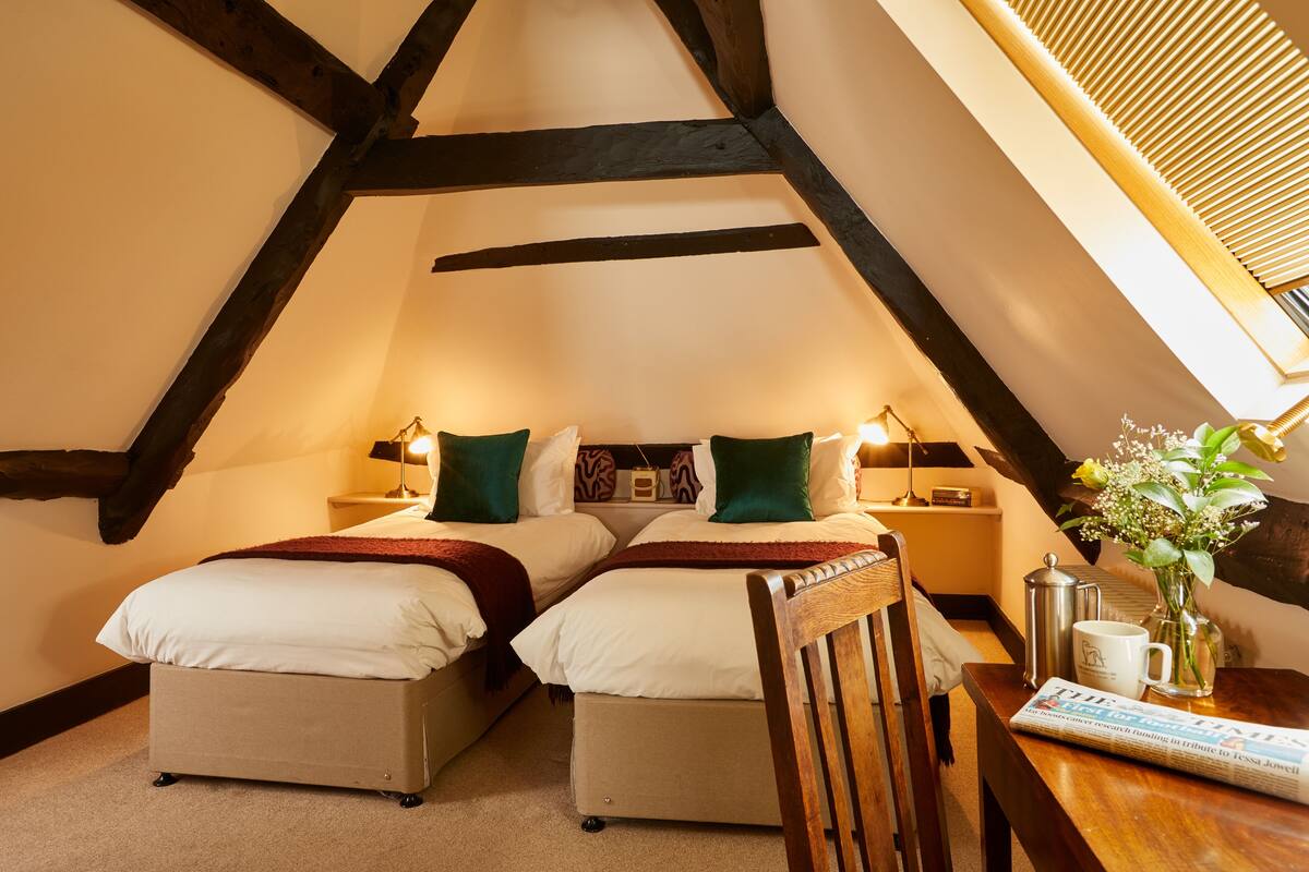 Um quarto de sótão acolhedor com duas camas, vigas de madeira, uma cadeira de madeira, uma mesa com um jornal e um vaso com flores.