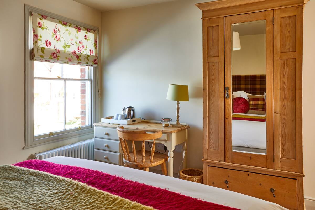 Quarto com cama, escrivaninha de madeira, cadeira, abajur, guarda-roupa e janela com cortina florida.
