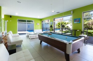 Sala com mesa de bilhar, sofás brancos e vista para o exterior.