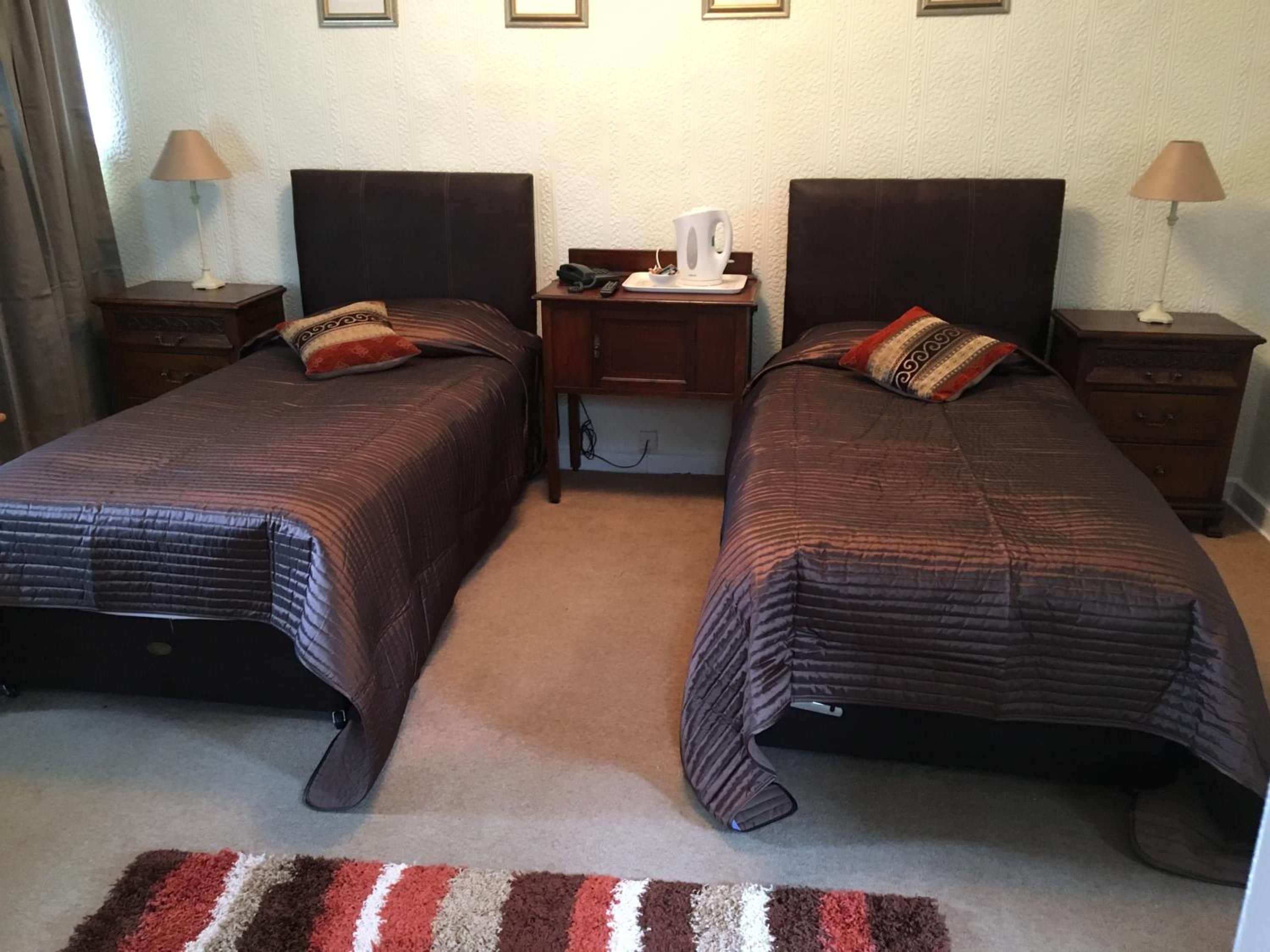 Quarto de hotel com duas camas de solteiro, uma mesinha com chaleira e criado-mudo com luminária.