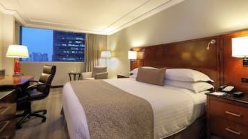 Quarto de hotel com uma cama grande, uma mesa com cadeira, mesa de cabeceira com telefone e vista da cidade ao entardecer.