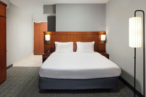 Quarto de hotel com uma cama grande, cabeceira de madeira, duas luminárias de cabeceira e uma mesinha de cabeceira.