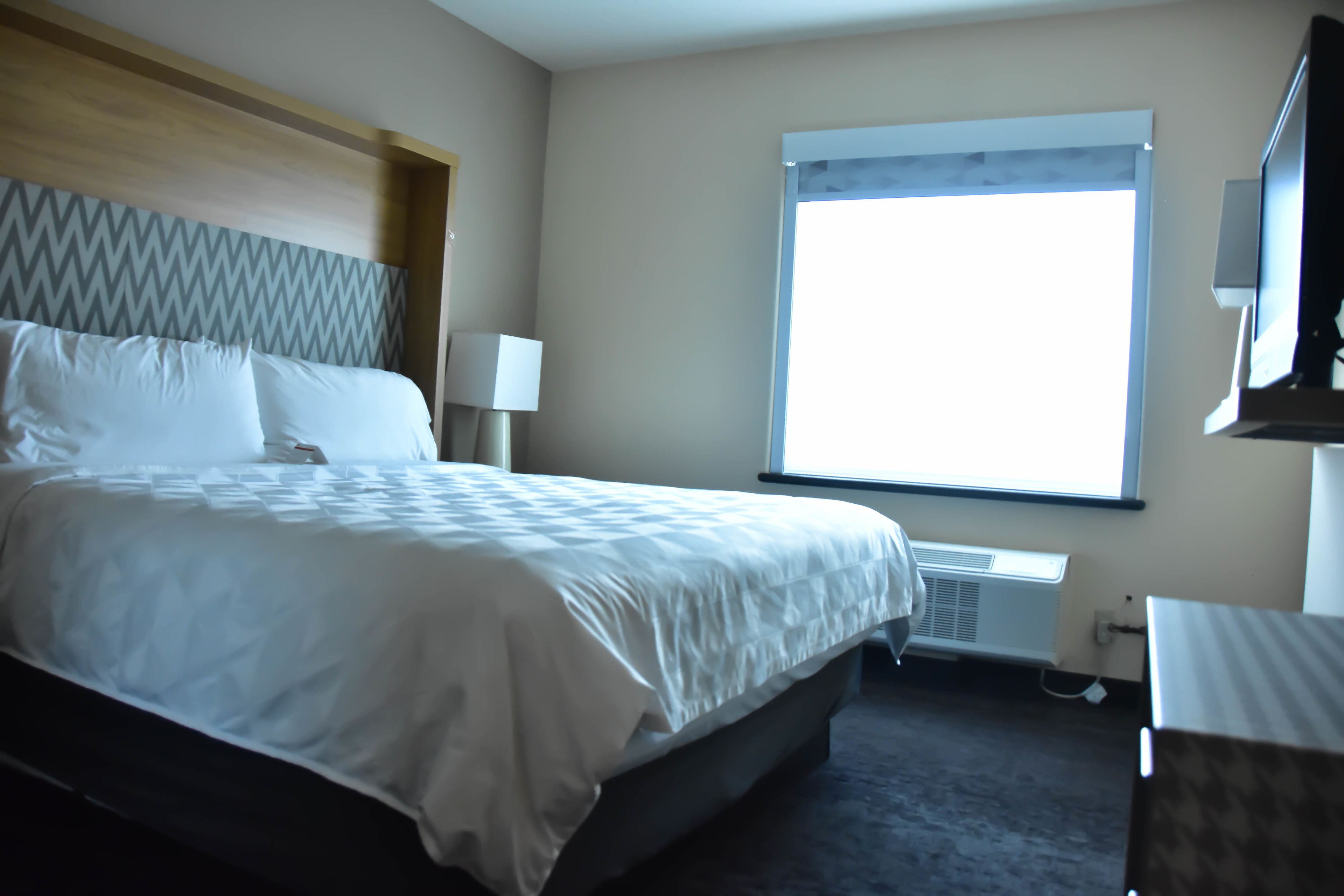 Quarto de hotel com uma cama grande, luminária de cabeceira, janela com persianas e televisão na parede.