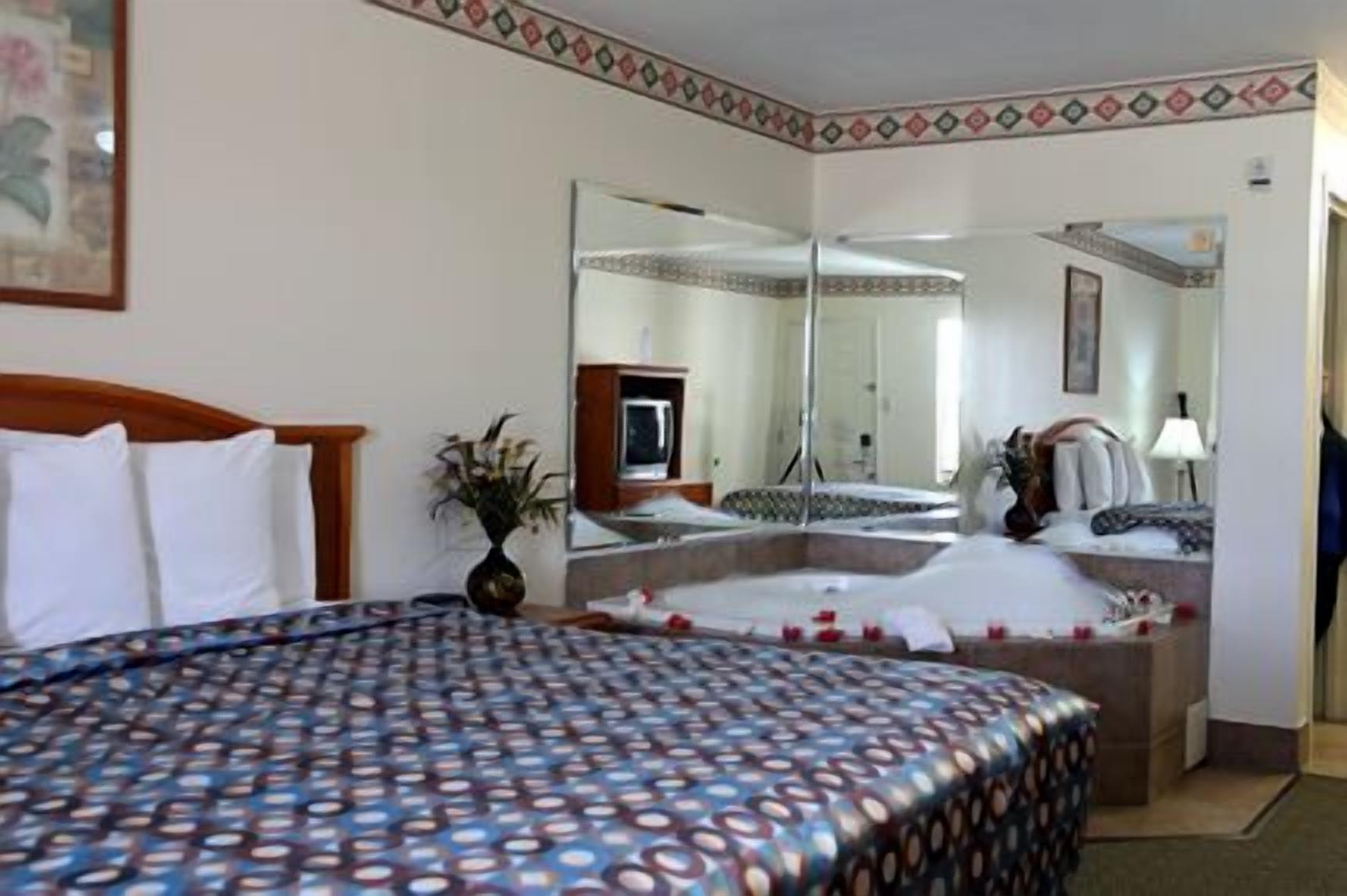 Quarto de hotel com uma cama, um espelho grande, uma televisão e uma pequena mesa com abajur.