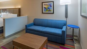Quarto de hotel com sofá azul, mesa de centro de madeira, mesa redonda pequena, luminária e tapete listrado colorido.