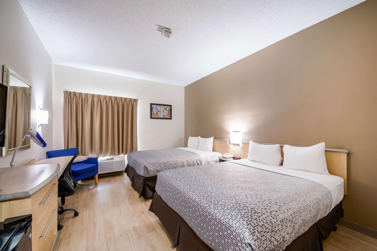 Quarto de hotel com duas camas, uma escrivaninha com cadeira, espelho, janela com cortinas e telefone fixado na parede.