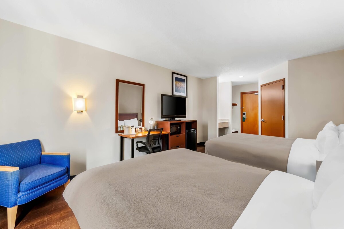 Quarto de hotel com duas camas, uma escrivaninha com computador e luminária, uma cadeira e uma televisão.