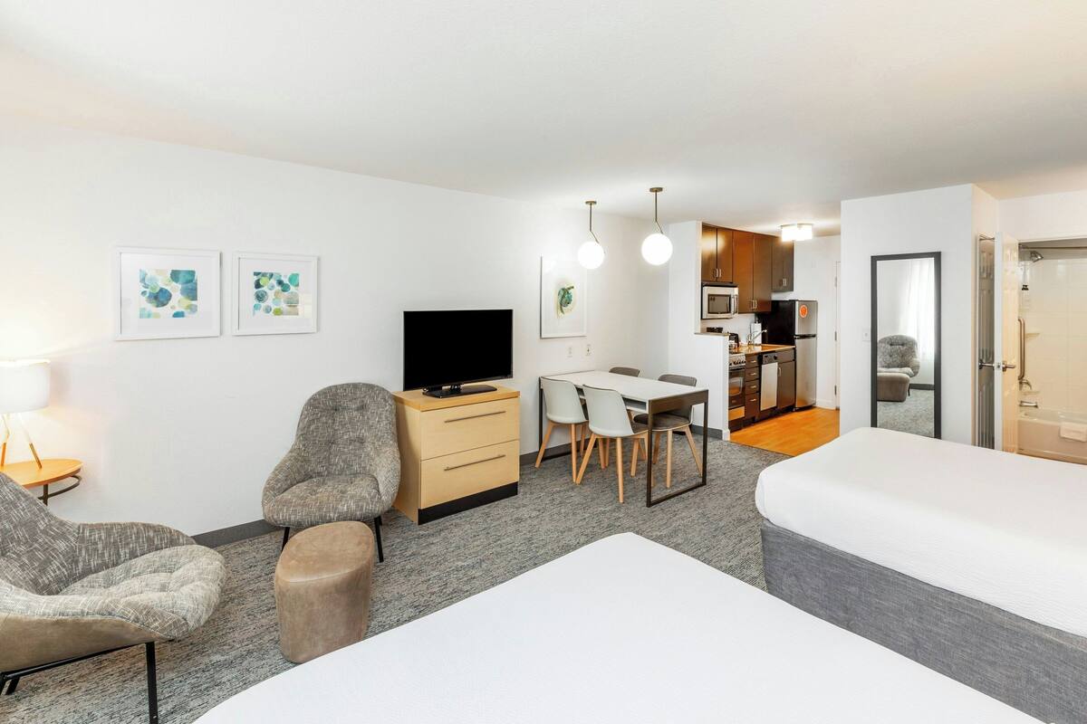 Quarto de hotel com cama, escrivaninha, cadeira, televisão e cozinha compacta.