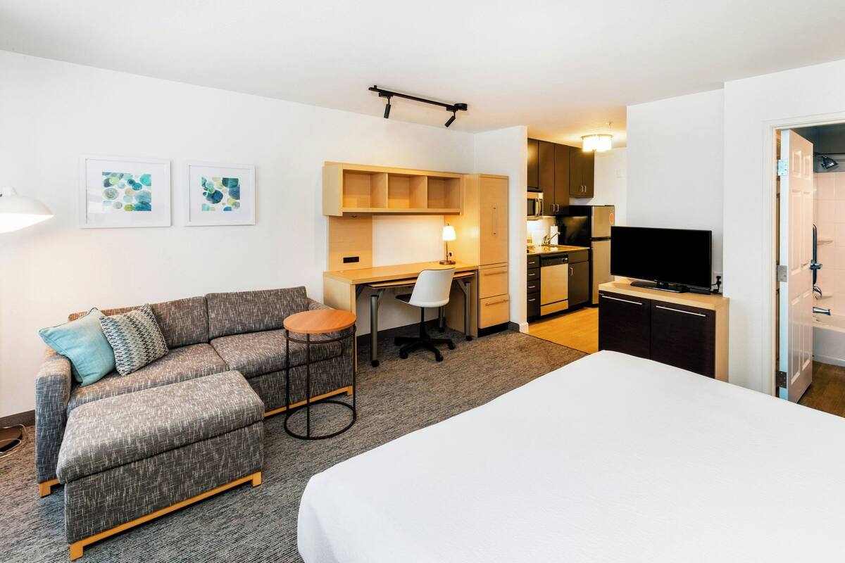 Quarto de hotel moderno com sofá, mesa redonda pequena, escrivaninha, televisão e cozinha compacta.