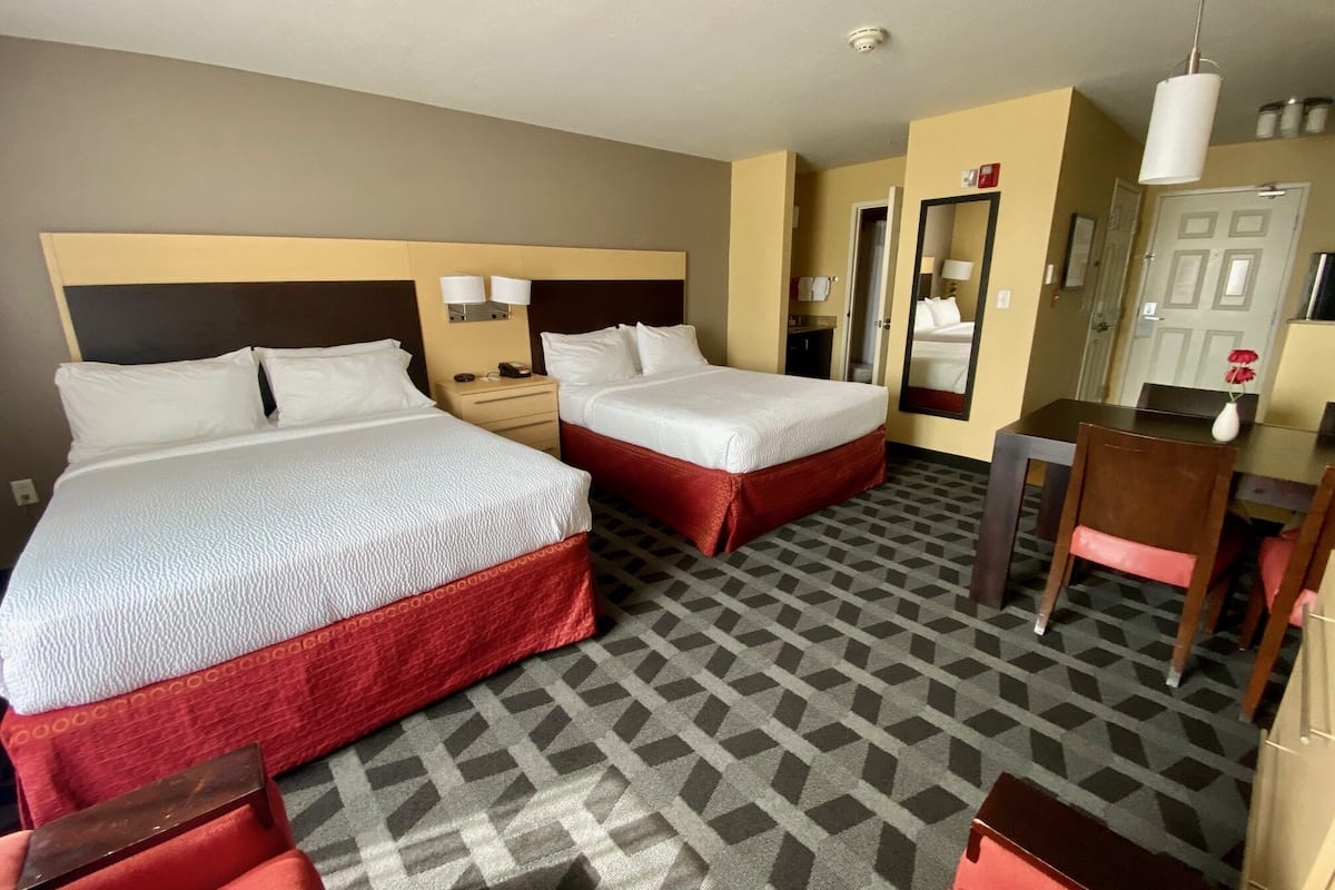 Quarto de hotel com duas camas, uma escrivaninha, uma cadeira, um espelho e um tapete estampado.