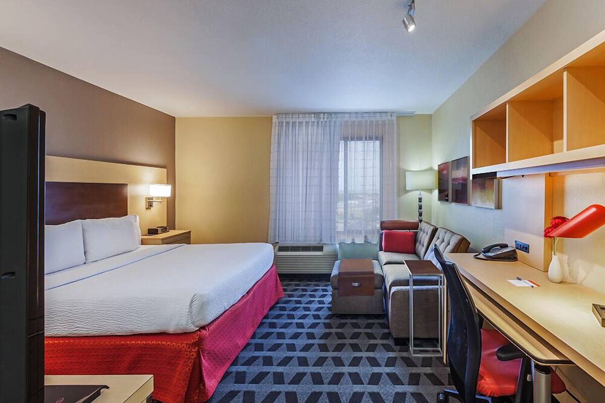 Quarto de hotel com cama, escrivaninha, cadeira, um pequeno sofá, televisão e janela com cortinas.