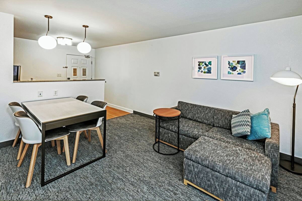 Uma sala de estar moderna com um sofá cinzento, uma mesa de centro redonda e uma área de jantar com mesa e cadeiras. Há duas obras de arte emolduradas na parede e um abajur.