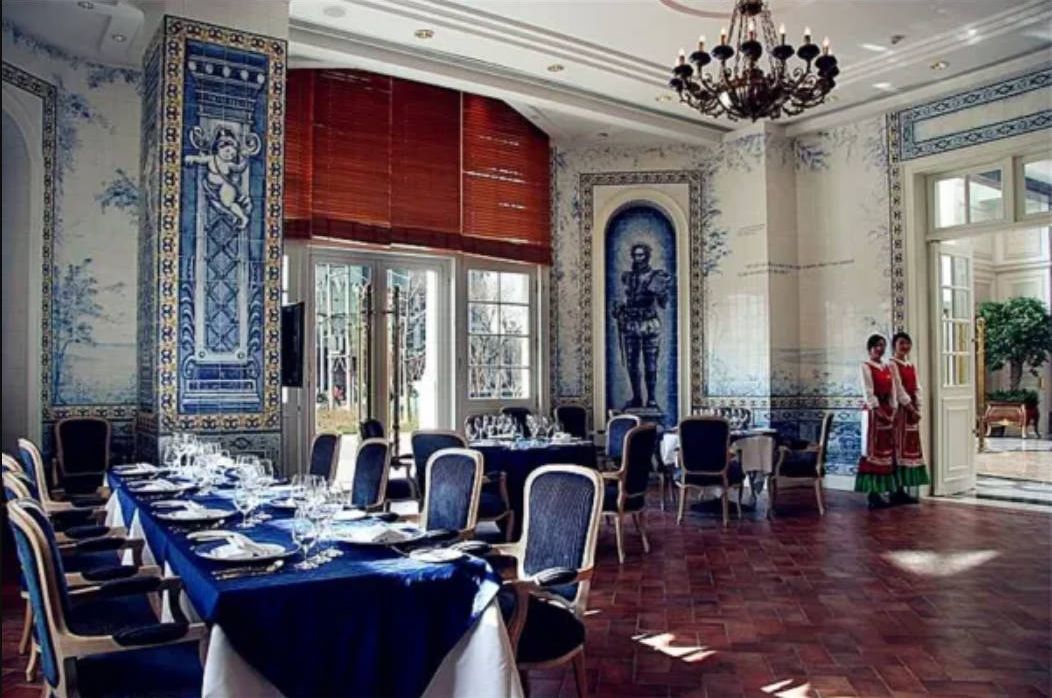 Uma sala de jantar formal com mesas postas para refeição, decorações azuis e brancas elaboradas nas paredes e um candelabro.