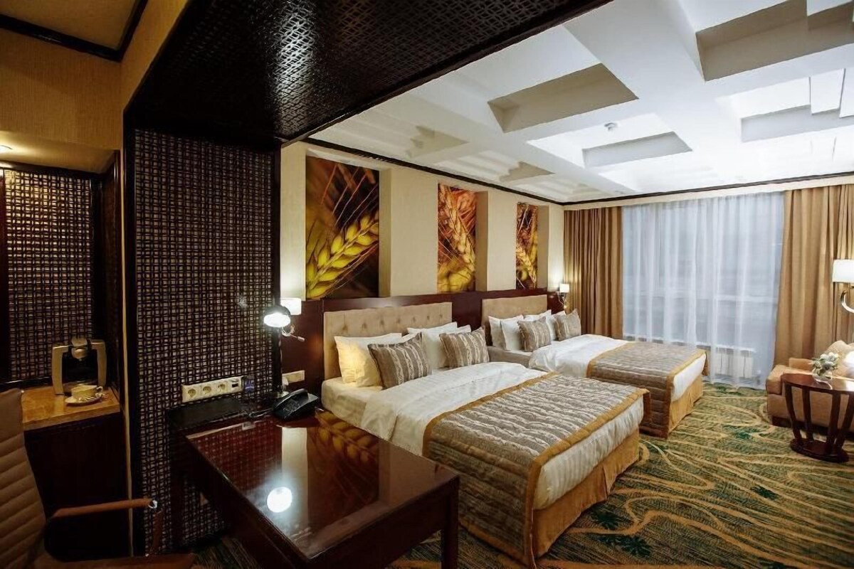 Quarto de hotel com duas camas, uma escrivaninha, uma cadeira, frigobar e uma janela grande com cortinas.