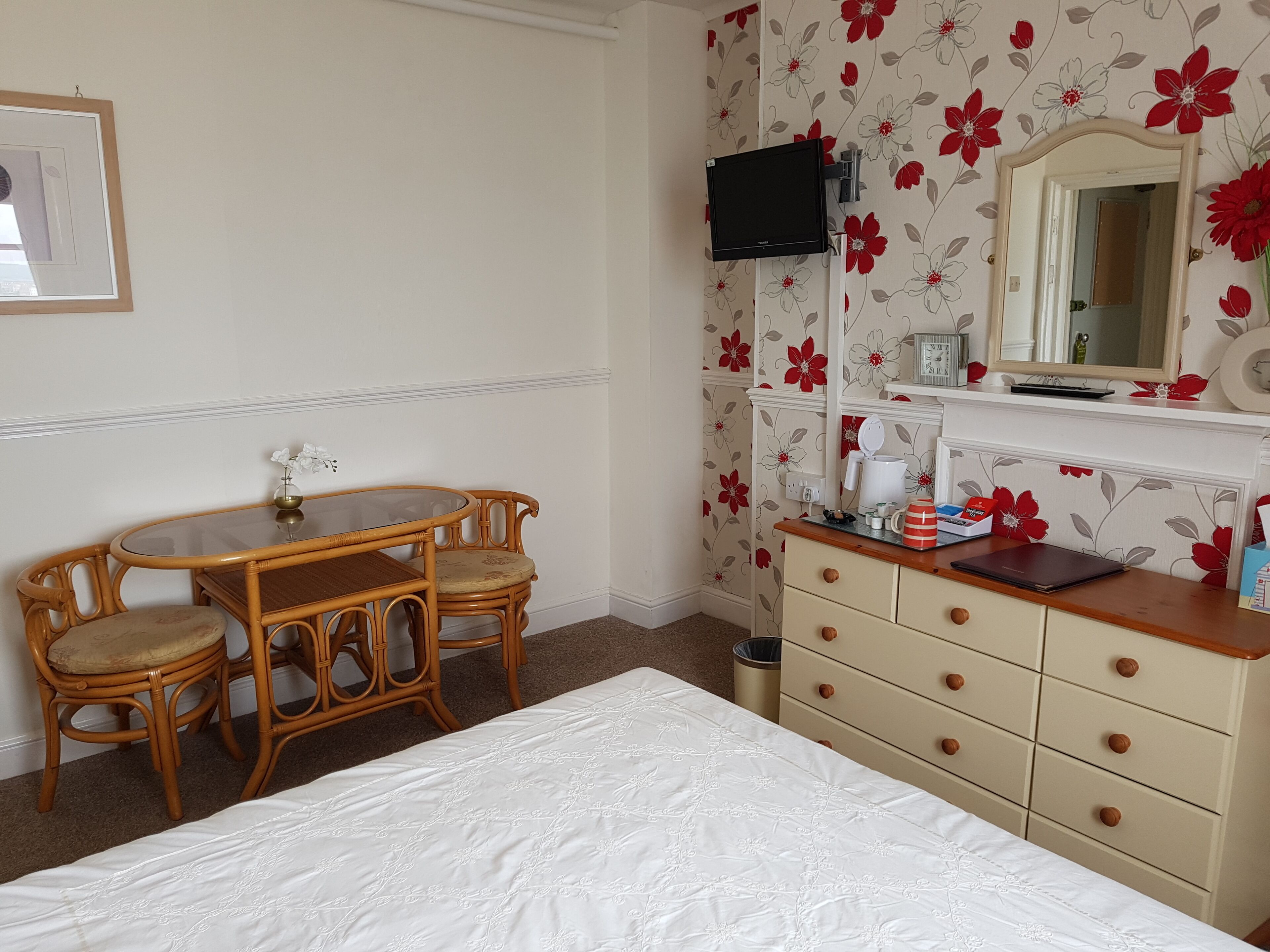 Quarto de hotel com uma cama, uma mesa de madeira com cadeiras, um aparador com gavetas, uma televisão fixada na parede e papel de parede floral.