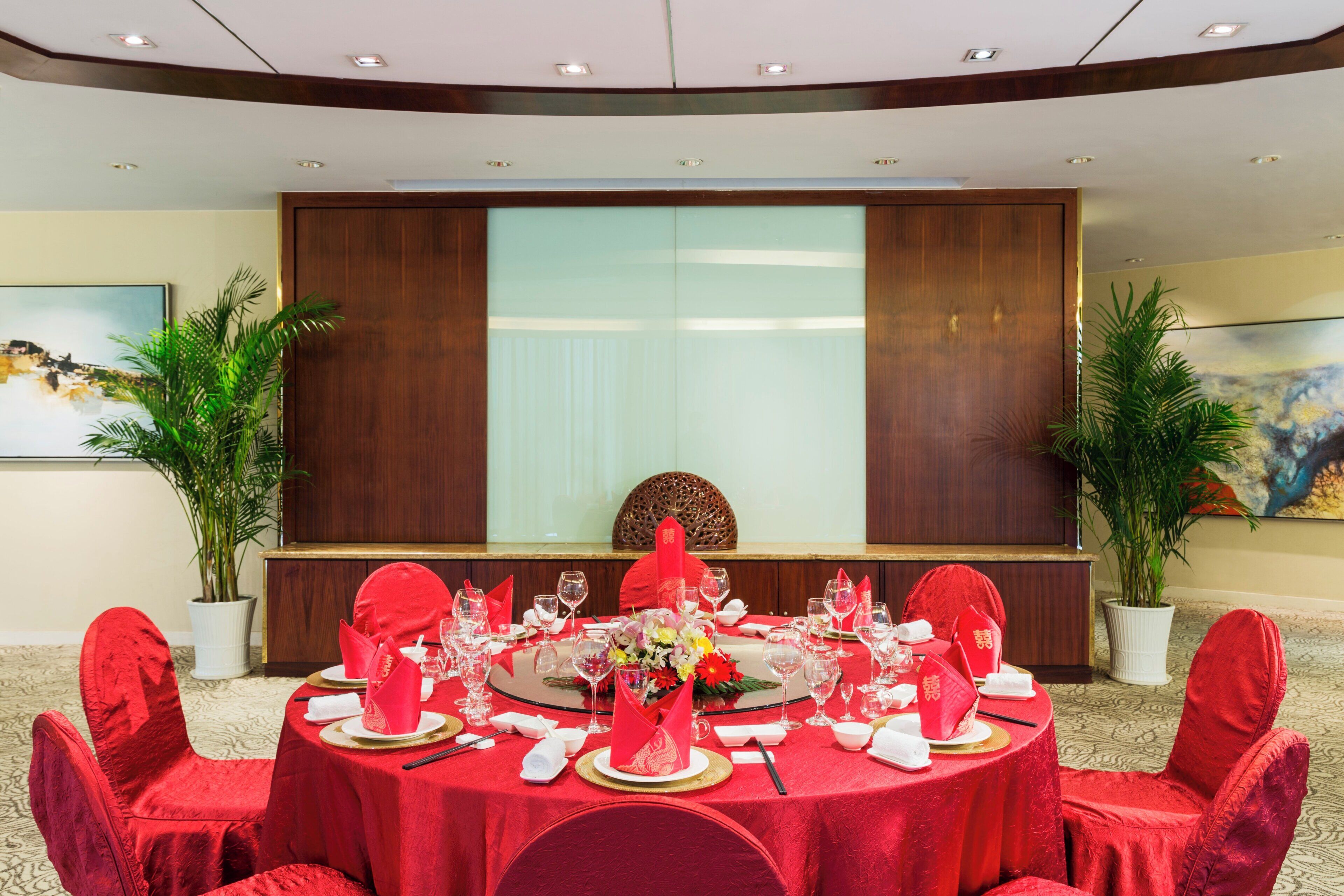 Sala de conferência com uma mesa redonda preparada para uma reunião, rodeada por cadeiras vermelhas, com um painel de madeira grande e plantas em vasos ao fundo.