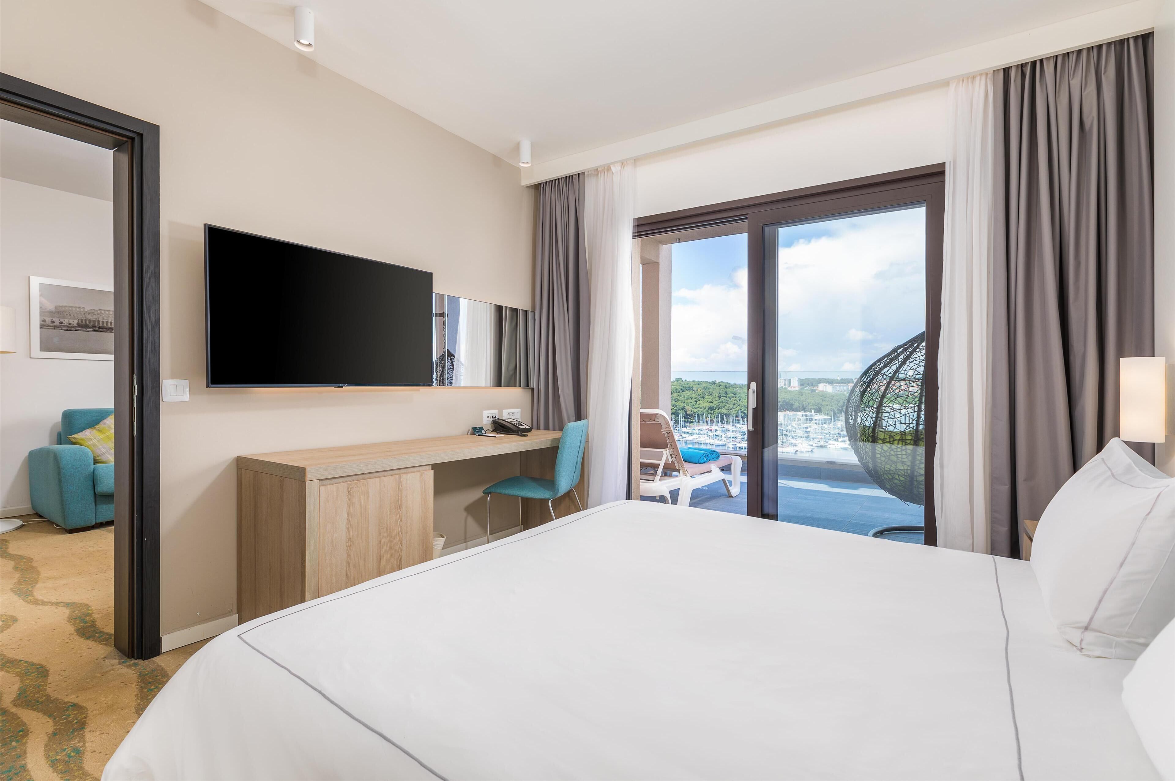 Quarto de hotel com uma cama grande, uma mesa de trabalho, televisão de tela plana, varanda com vista para a cidade e uma ponte, e uma cadeira.