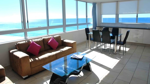 Quarto com uma grande janela com vista para o oceano, um sofá marrom com almofadas cor-de-rosa, uma mesa de centro de vidro e uma área de jantar com cadeiras e uma mesa.