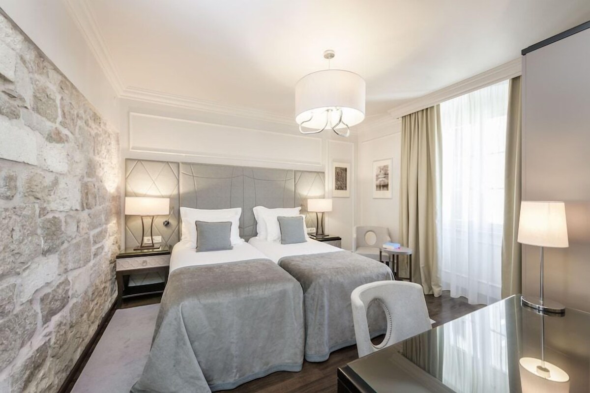 Quarto de hotel com duas camas, parede de pedra, uma janela grande com cortinas e uma mesa com abajur.