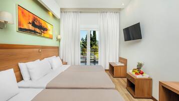 Quarto de hotel com uma cama grande, televisão, cesta de frutas e vista para árvores pela janela.