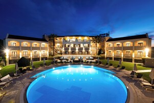 Um hotel com uma grande piscina cercada por espreguiçadeiras e guarda-sóis, fachada de edifício bem iluminada e céu claro.