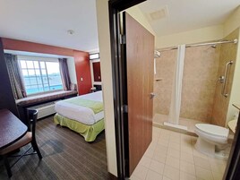 Quarto de hotel com cama, escrivaninha, janela com vista, chuveiro e vaso sanitário.