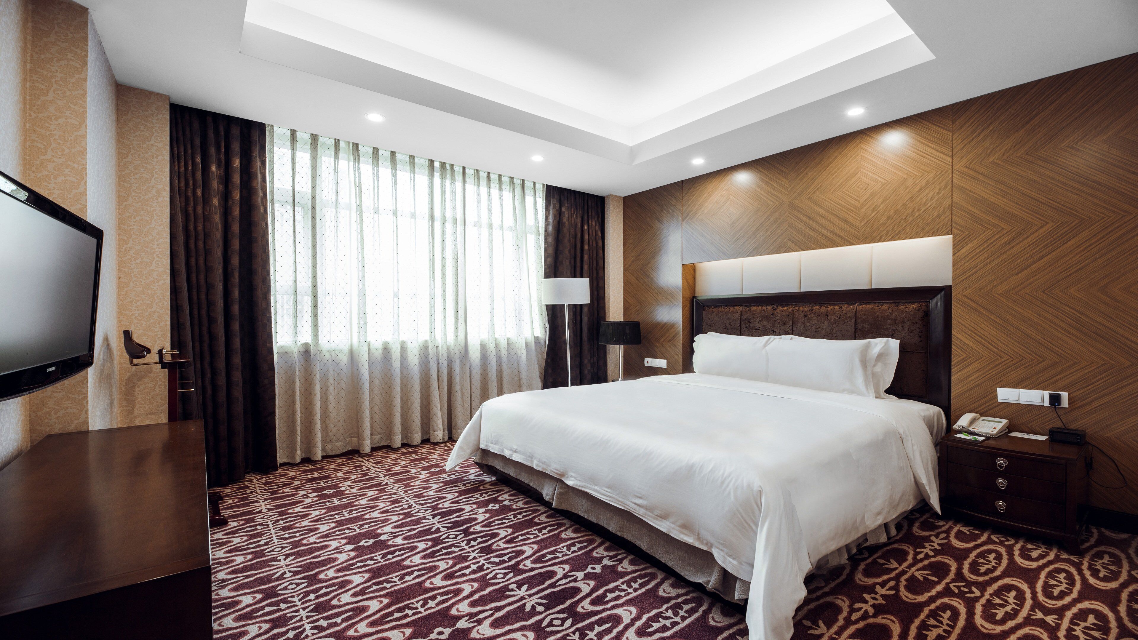 Quarto de hotel com uma cama grande, televisão, secretária e carpete estampado.