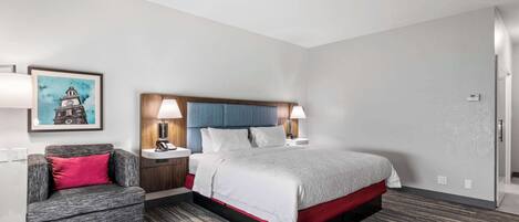 Quarto de hotel com cama, cadeira, mesa de cabeceira e um quadro na parede.