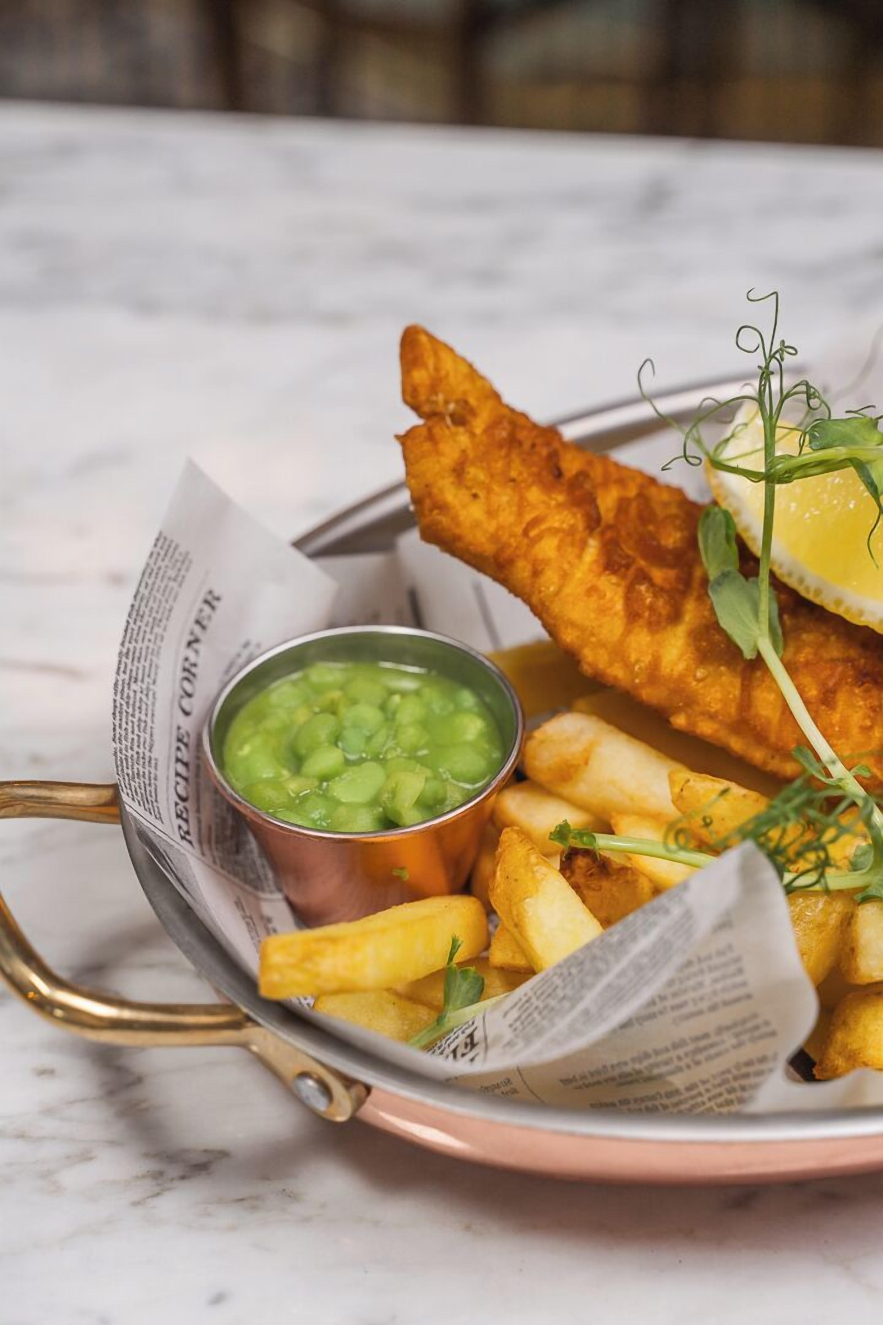 Uma cesta com peixe frito e batatas, acompanhada de ervilhas amassadas e um limão.