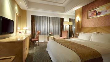 Quarto de hotel com uma cama grande, uma escrivaninha com televisão, uma cadeira, uma mesinha com um vaso de flores e uma janela grande com cortinas.