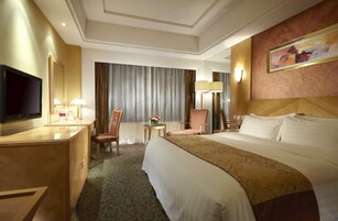 Quarto de hotel com uma cama grande, uma escrivaninha com televisão, uma cadeira, uma mesinha com um vaso de flores e uma janela grande com cortinas.
