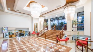 Um hall amplo com piso estampado em padrão geométrico, escada e áreas de estar com assentos vermelhos.