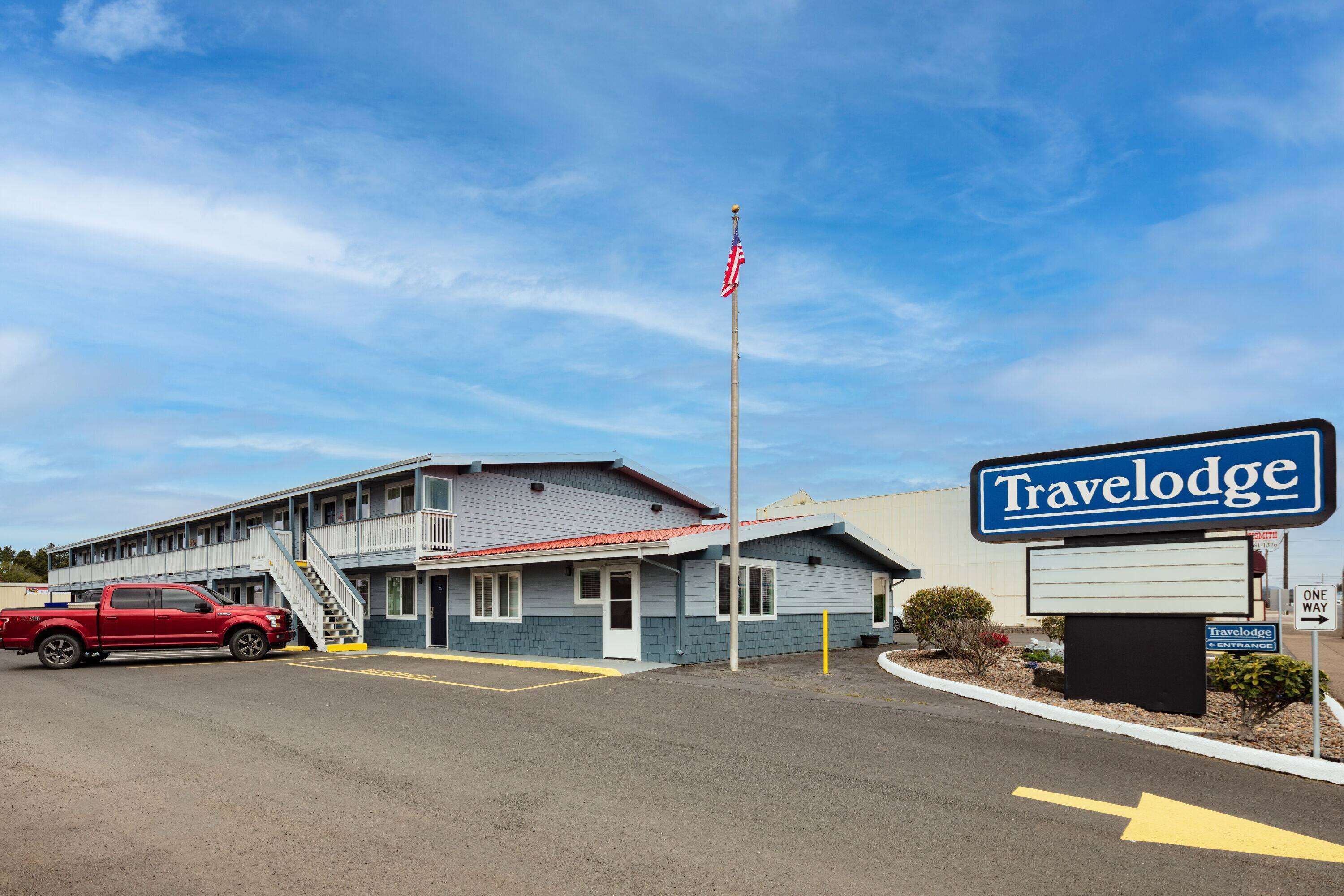Um motel Travelodge com uma caminhonete vermelha estacionada em frente, um mastro com a bandeira americana e uma placa indicando entrada em sentido único.